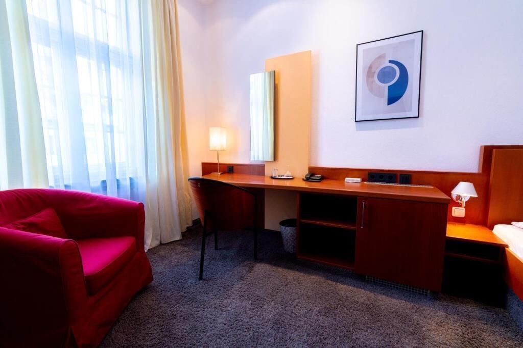 Quarto de hotel com escrivaninha, uma cadeira vermelha, cama, mesa de cabeceira, luminária, lixeira e uma obra de arte emoldurada na parede.