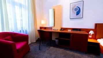 Quarto de hotel com escrivaninha, uma cadeira vermelha, cama, mesa de cabeceira, luminária, lixeira e uma obra de arte emoldurada na parede.