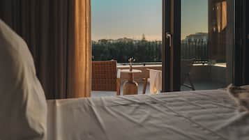 Quarto de hotel com cama, uma pequena mesa redonda, uma cadeira e vista para uma varanda com grades e árvores.