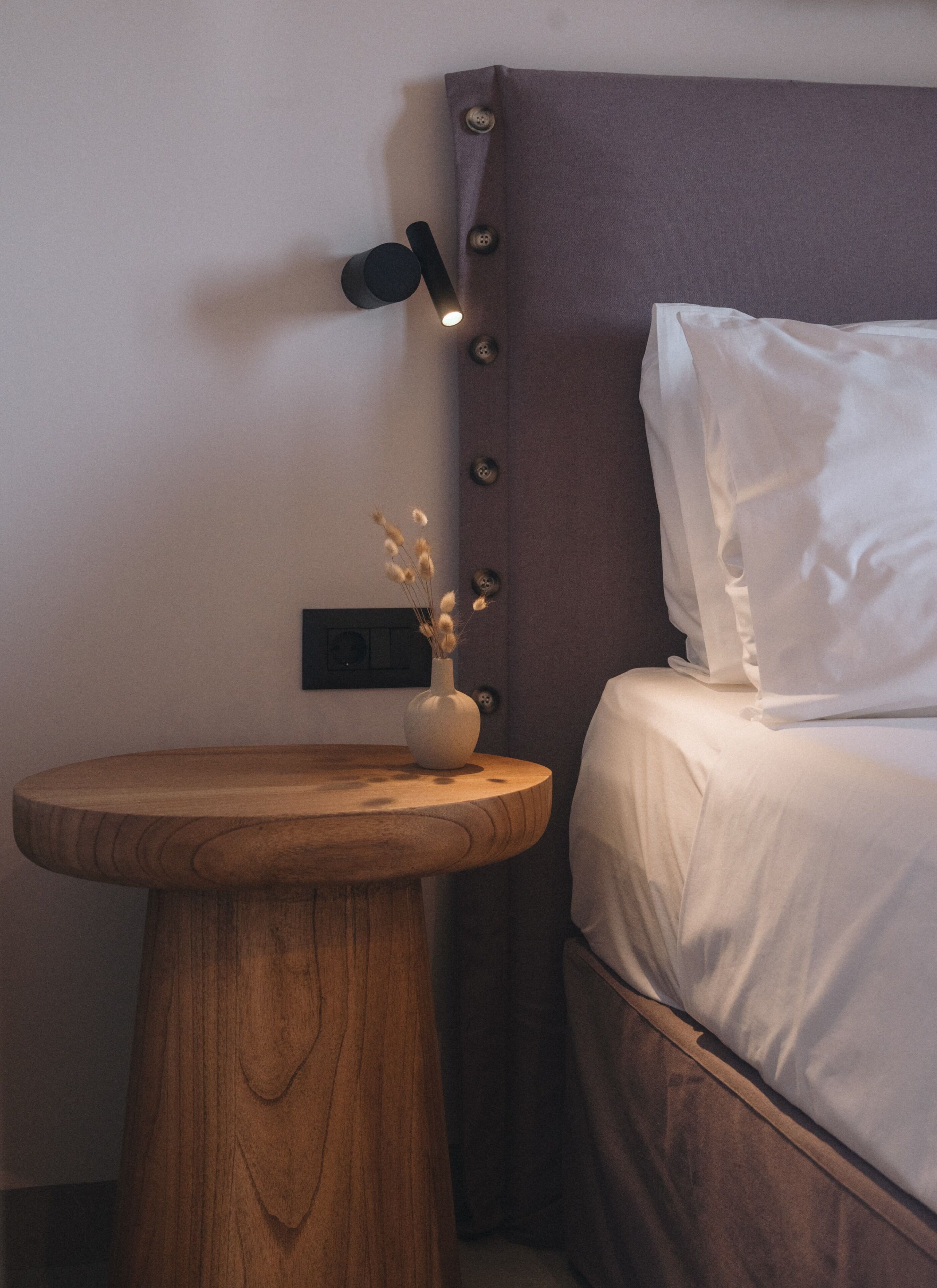 Uma cama bem arrumada com roupa de cama branca, um criado-mudo de madeira redondo com um vaso de flores secas e um aplique na parede.