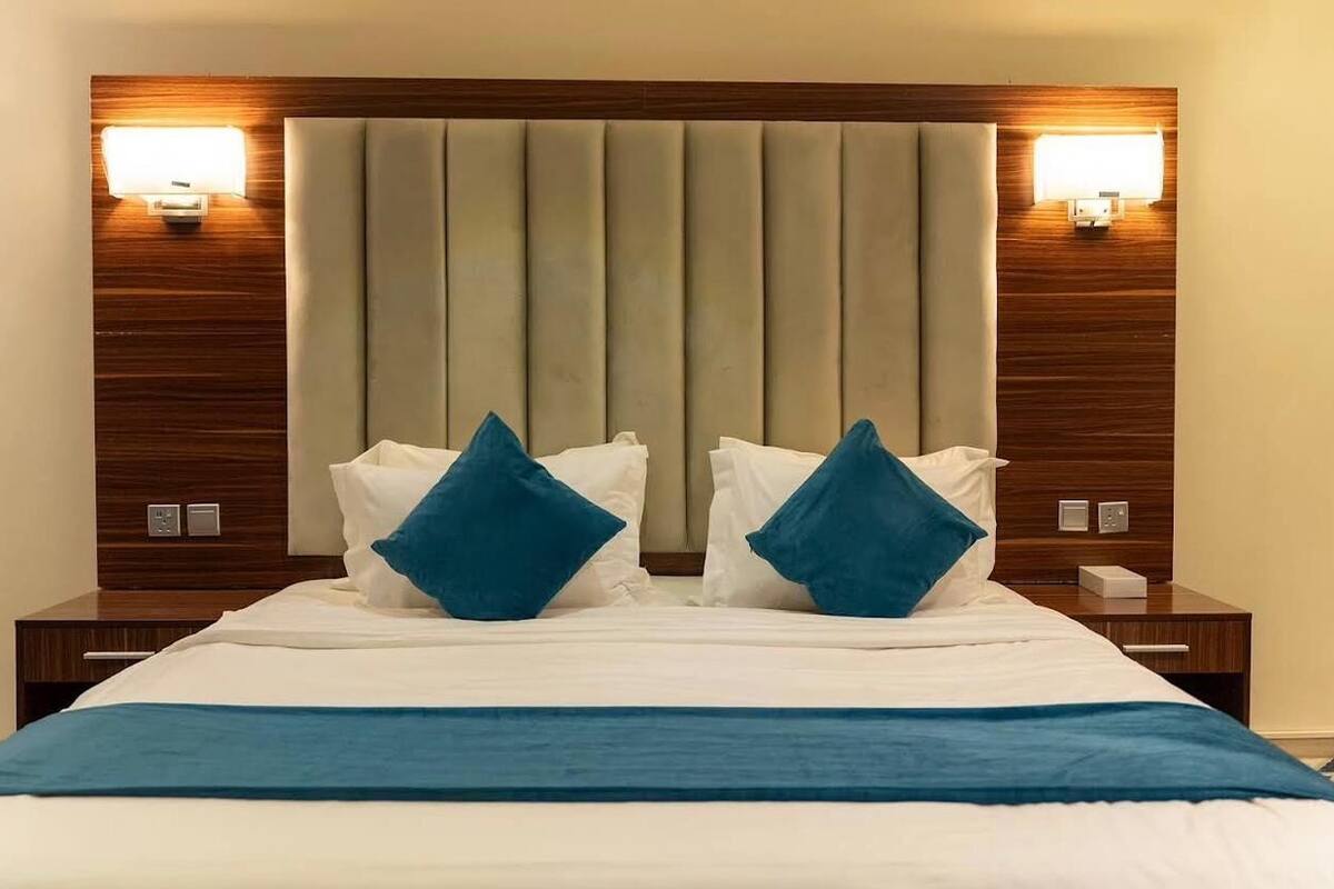 Quarto de hotel com uma cama grande, duas mesas de cabeceira, cabeceira com linhas verticais e luminárias fixadas na parede.