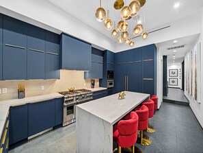 Uma cozinha moderna com armários azul-escuros, uma ilha de mármore, banquetas vermelhas e um lustre esférico singular.