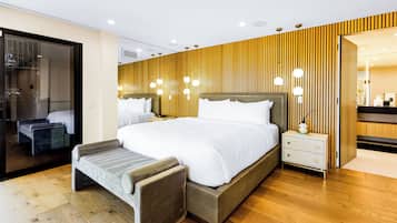 Quarto de hotel moderno com uma cama grande, mesas de cabeceira, um banco e vista para a cidade.