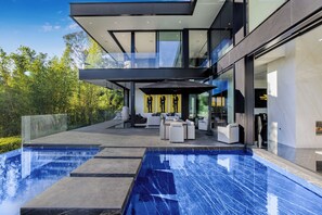 Uma área moderna de piscina ao ar livre, com um deque envidraçado, assentos externos e vista para o verde circundante.