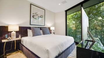 Quarto moderno com uma cama grande, abajures, uma obra de arte emoldurada e vista para o verde através de uma janela ampla.