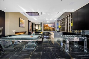 Uma área de piscina moderna com mesa de bilhar em recinto de vidro, espaço de estar com sofá e cadeiras, e um grande mural com paisagem na parede.