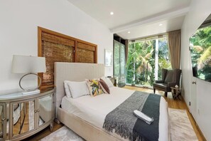 Quarto moderno com uma cama grande, mesa de cabeceira, luminária, cadeira e vista para o verde através de amplas janelas.