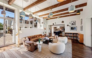 Uma sala de estar ampla com um sofá de couro, um banquinho branco e uma mesa de centro. Há uma estante cheia de livros e uma área de refeições ao fundo.