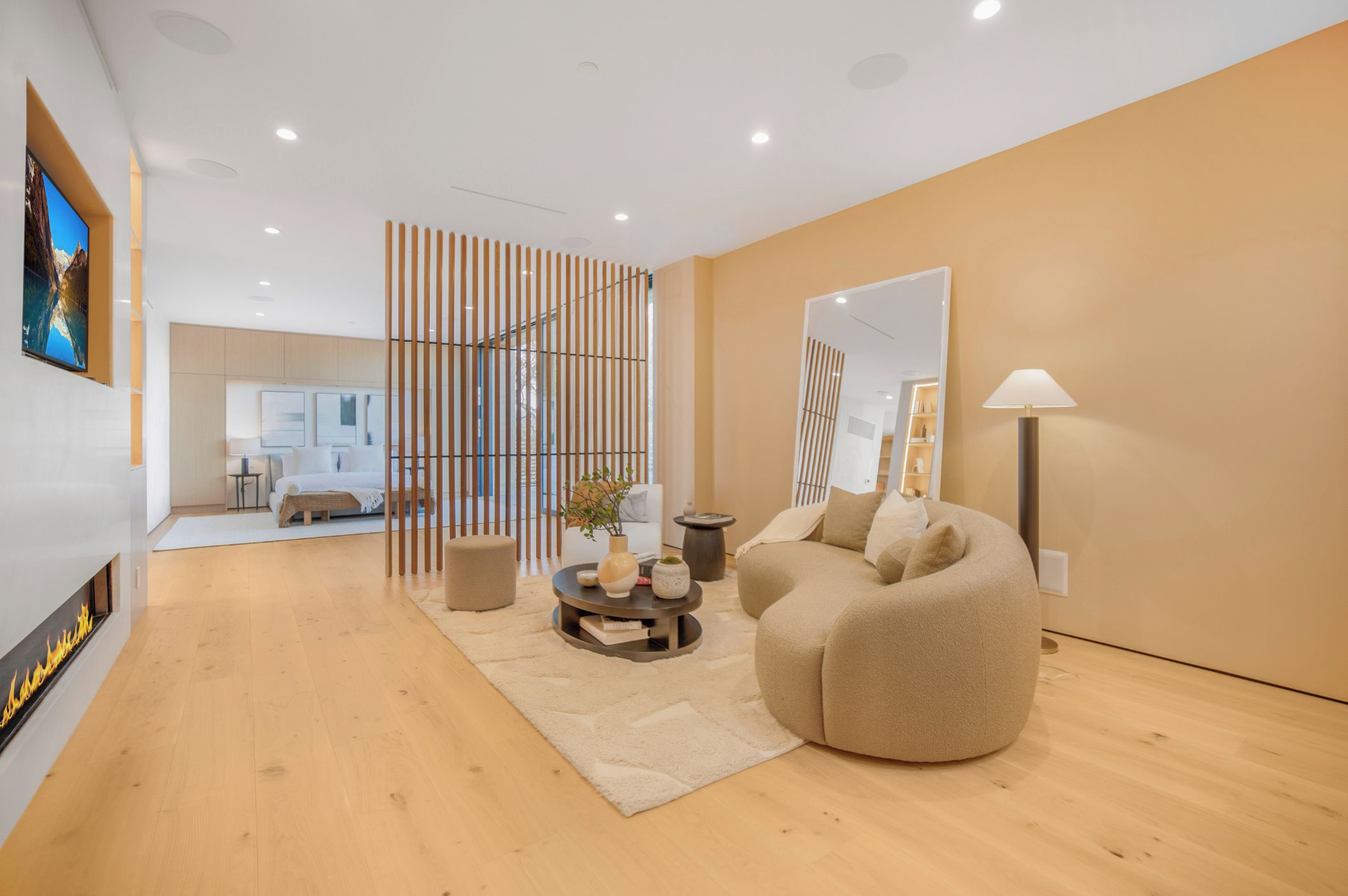 Uma sala de estar moderna com um sofá bege, uma mesa de centro redonda com objetos decorativos, uma lareira e um espelho grande na parede.