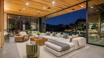 Uma sala de estar moderna com um sofá seccional grande, uma mesa de centro e uma poltrona verde. Há uma parede de vidro que oferece vista para uma piscina e árvores.
