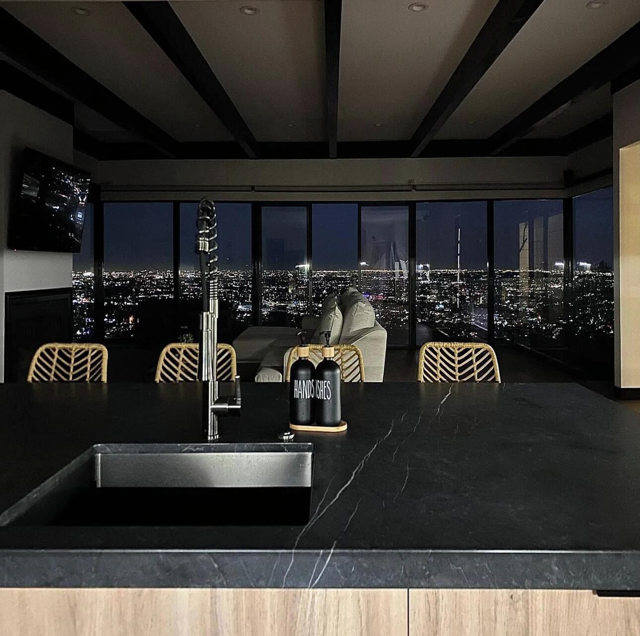 Uma cozinha moderna com bancada preta, uma pia e duas garrafas pretas com a etiqueta "HANDS SIEGE". Há uma janela grande com vista para a cidade à noite.