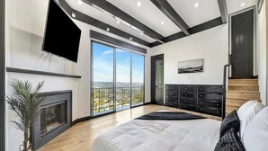 Quarto com cama grande, lareira, TV na parede e varanda com vista para a cidade.