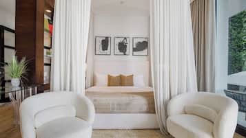 Quarto com cama, duas cadeiras, mesa de cabeceira, planta em vaso e obra de arte na parede.