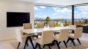 Uma área de refeições moderna com uma mesa grande e seis cadeiras, uma televisão de tela plana na parede e vista para a cidade através de amplas janelas.