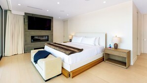 Quarto de hotel moderno com uma cama grande, televisão de tela plana fixada em parede de pedra, mesa de cabeceira com abajur e um pequeno espaço de estar com uma poltrona e um cobertor.