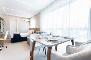Quarto de hotel moderno com área de refeições, cama e uma janela grande com cortinas leves.