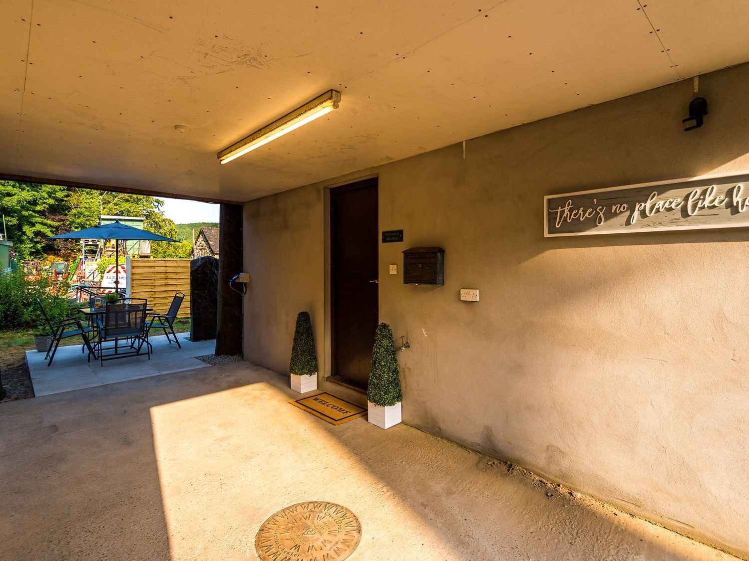 Uma área externa coberta com mesa e cadeiras, um tapete de boas-vindas e uma placa na parede.