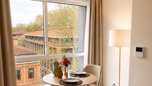 Quarto moderno com mesa de jantar posta para dois, luminária de chão e vista para edifícios através de grandes janelas.