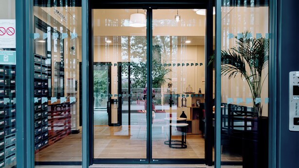 Entrada de hotel com portas de vidro, balcão de recepção e área de estar com uma planta.