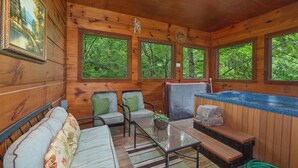 Interior de uma cabana com sofá, cadeiras, mesa, hidromassagem e janelas amplas com vista para uma floresta.