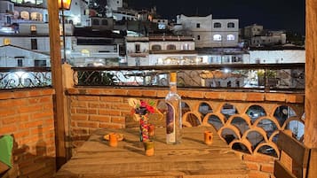 Uma mesa de madeira com cadeiras e uma garrafa, com o horizonte urbano iluminado ao fundo à noite.