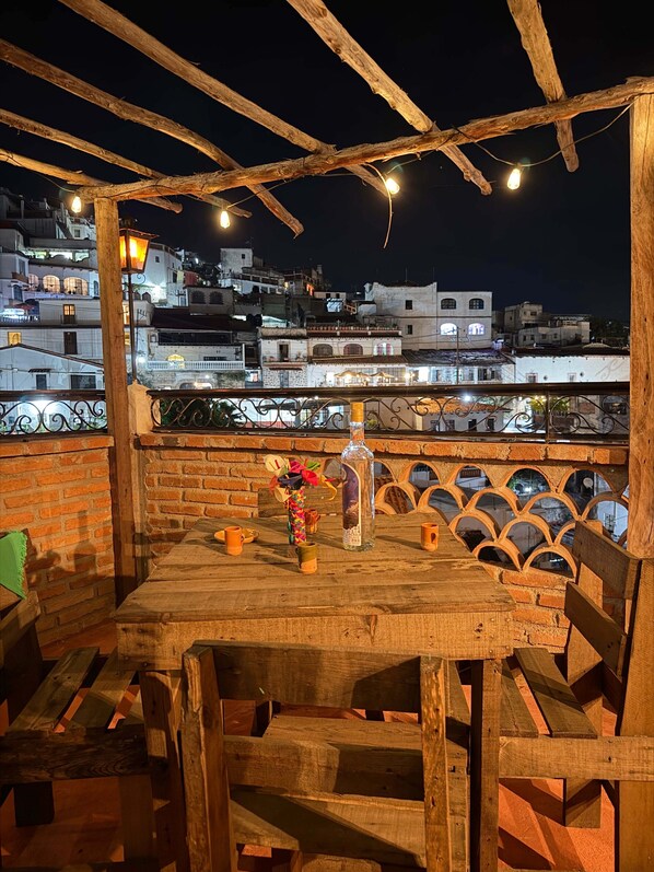 Uma mesa de madeira com cadeiras e uma garrafa, com o horizonte urbano iluminado ao fundo à noite.
