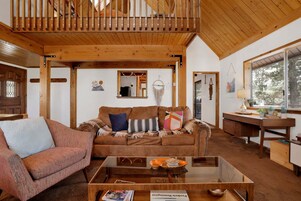 Uma sala de estar aconchegante com teto de madeira, um sofá marrom, uma mesa de centro de vidro e uma escrivaninha com abajur.