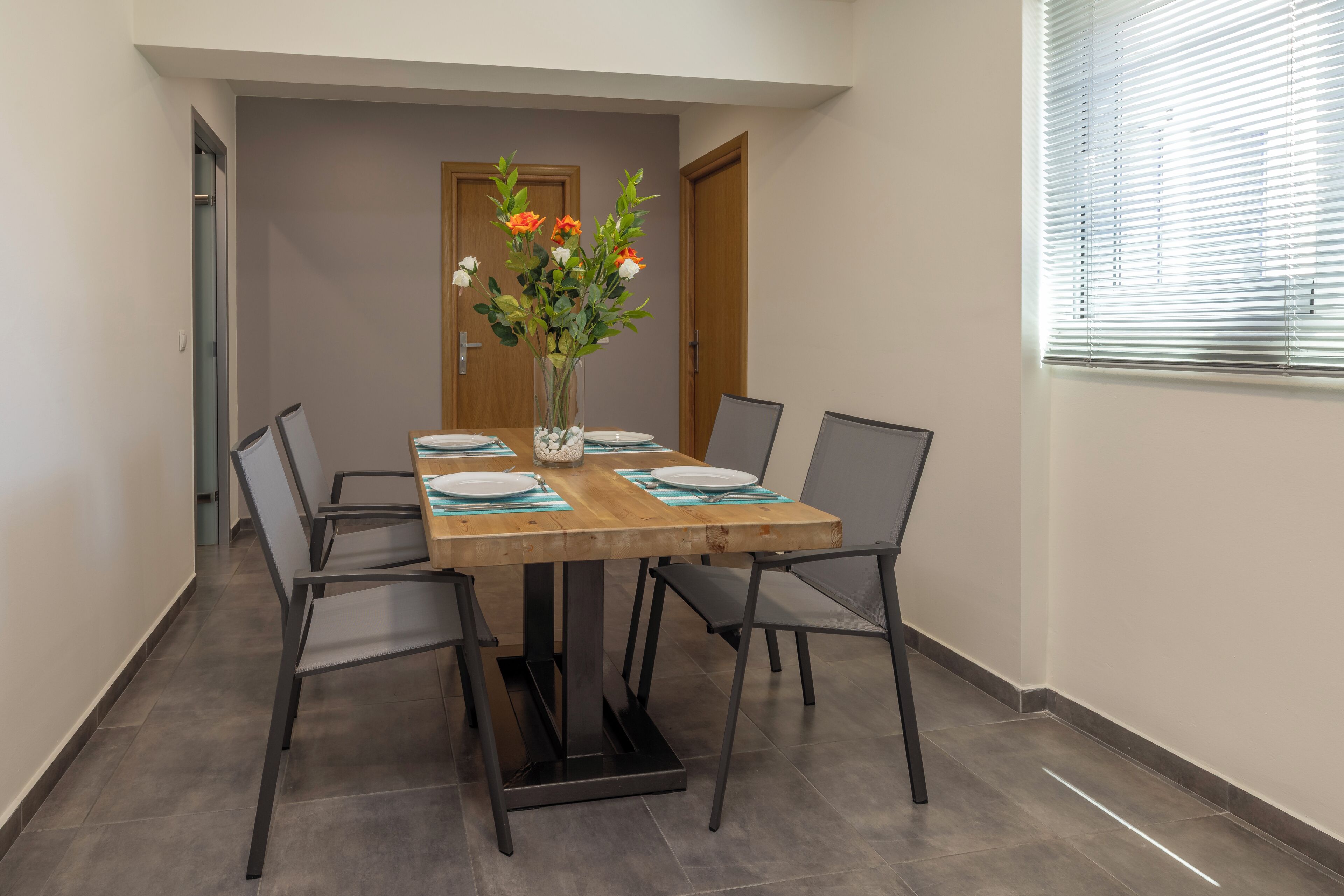Uma sala de jantar com uma mesa de madeira, seis cadeiras, um vaso com flores e uma porta ao fundo.