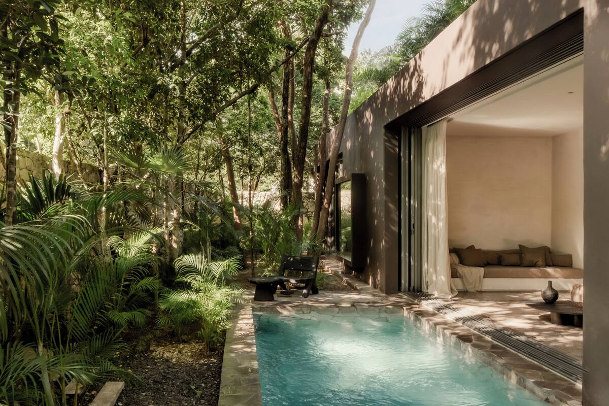 Piscina rodeada por vegetação exuberante, área de estar com sofá e mesa, e um edifício moderno com grandes janelas.