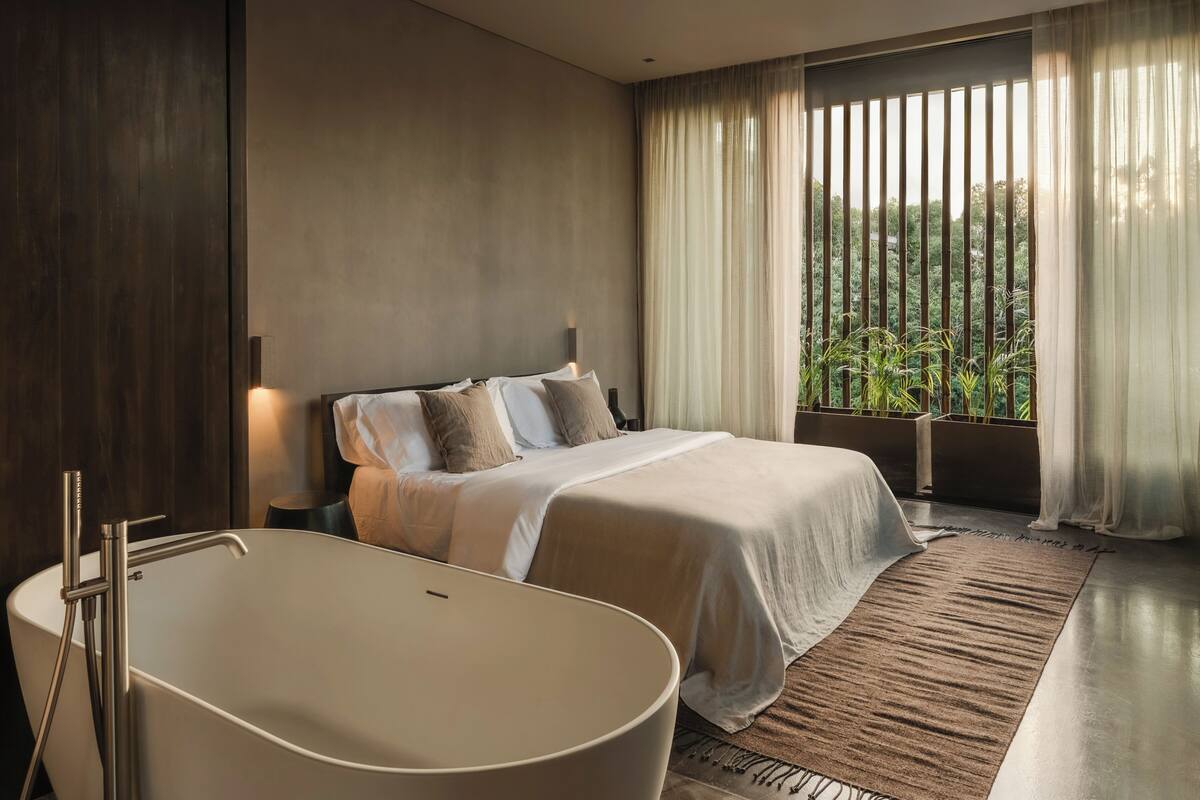 Quarto moderno com uma banheira grande, cama com várias almofadas, mesa de cabeceira e janela com cortinas.