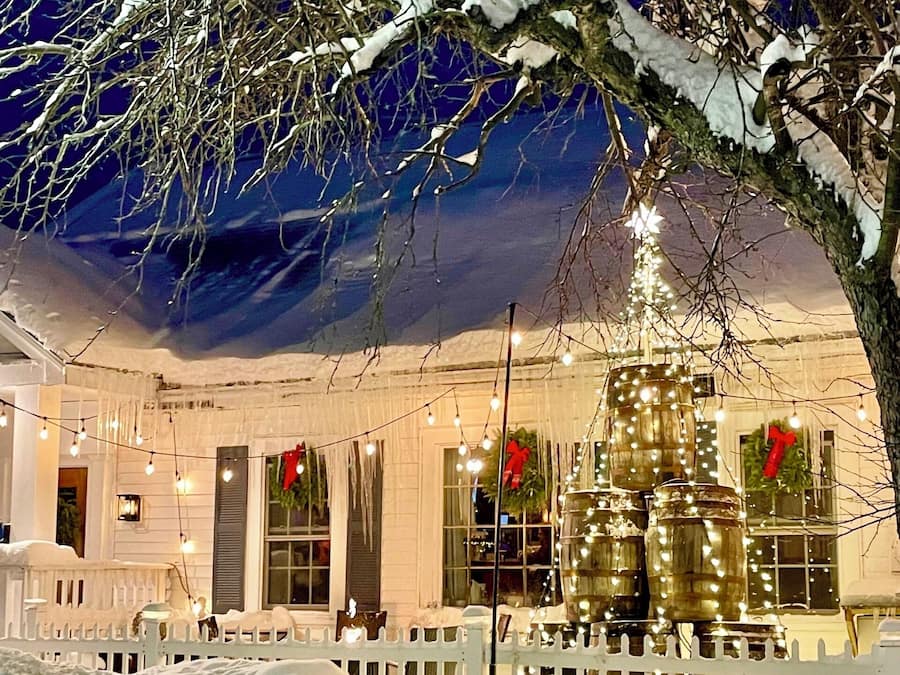 Área externa coberta de neve com cerca branca, árvore com galhos carregados de neve e um edifício com janelas decoradas com luzes e guirlandas.