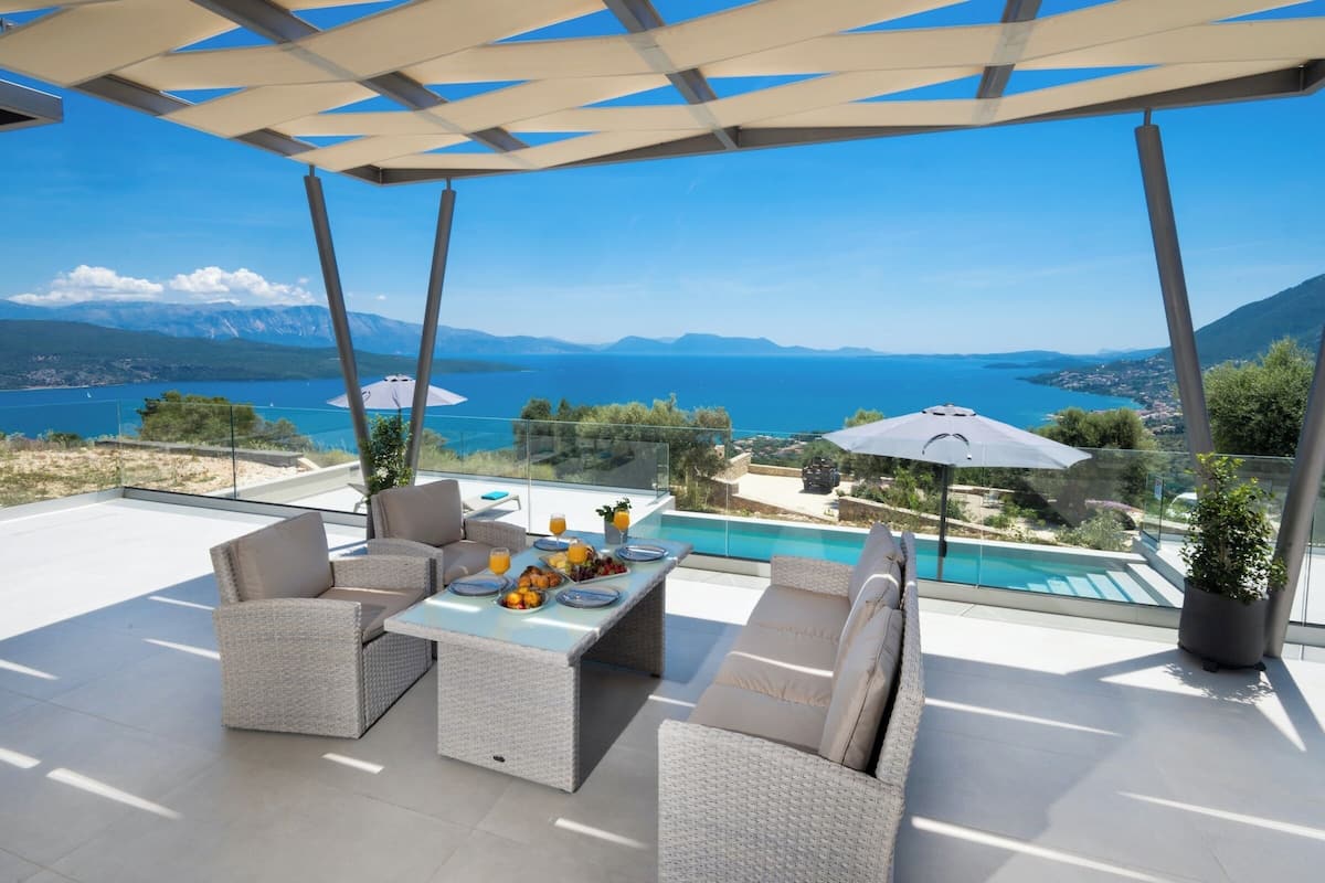 Um terraço com mobiliário de vime, uma mesa com frutas e bebidas, e uma piscina com guarda-sóis.