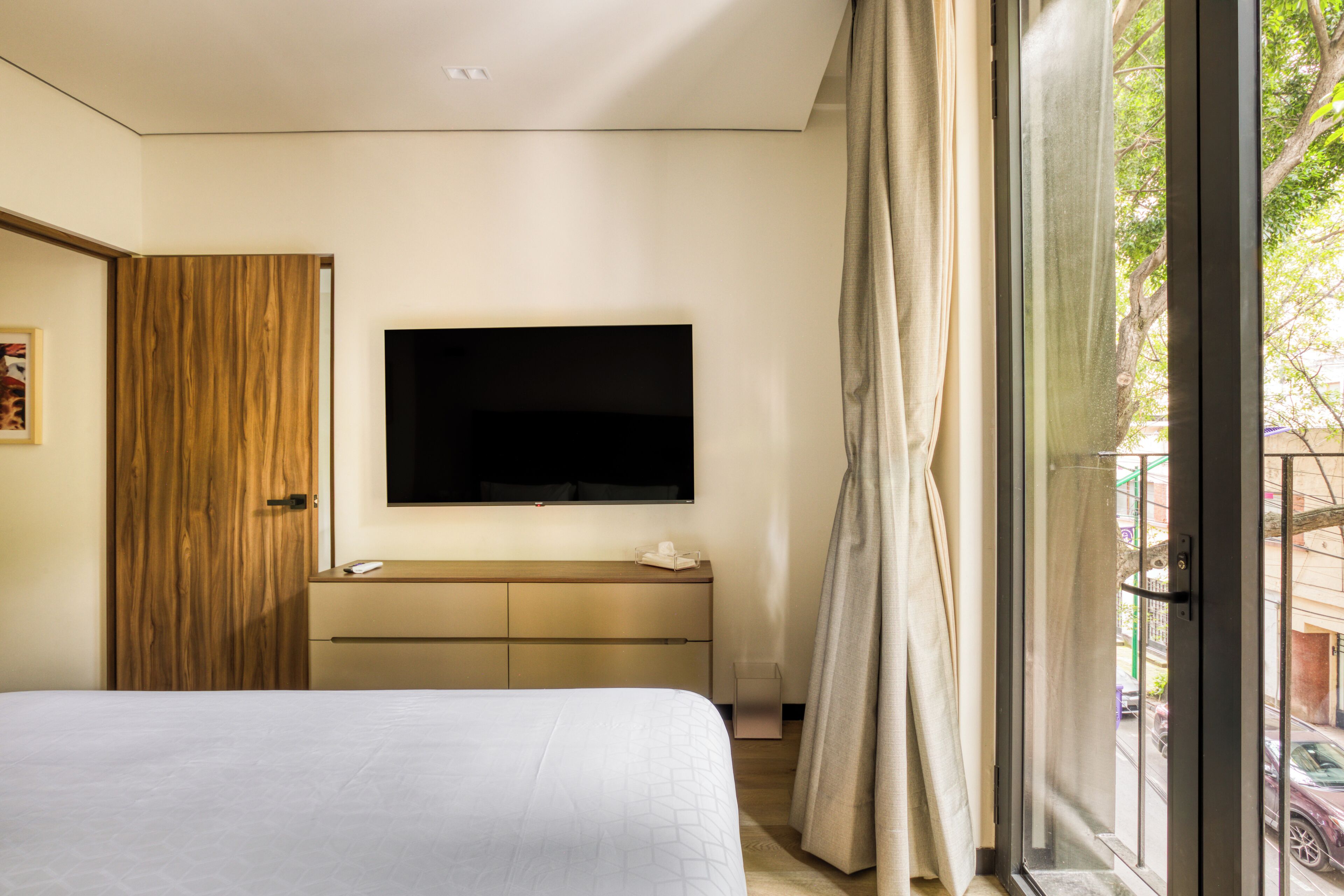 Quarto com cama, televisão na parede, guarda-roupa de madeira e janela com cortinas.