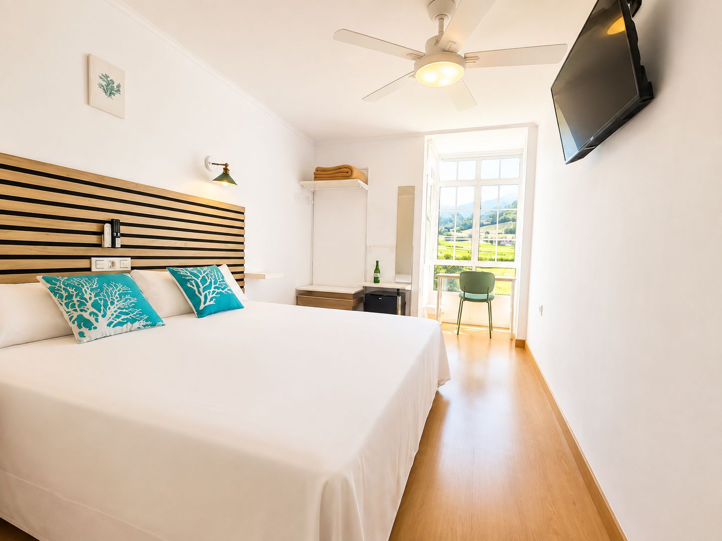 Quarto com cama, escrivaninha, cadeira, televisão e vista para o exterior.