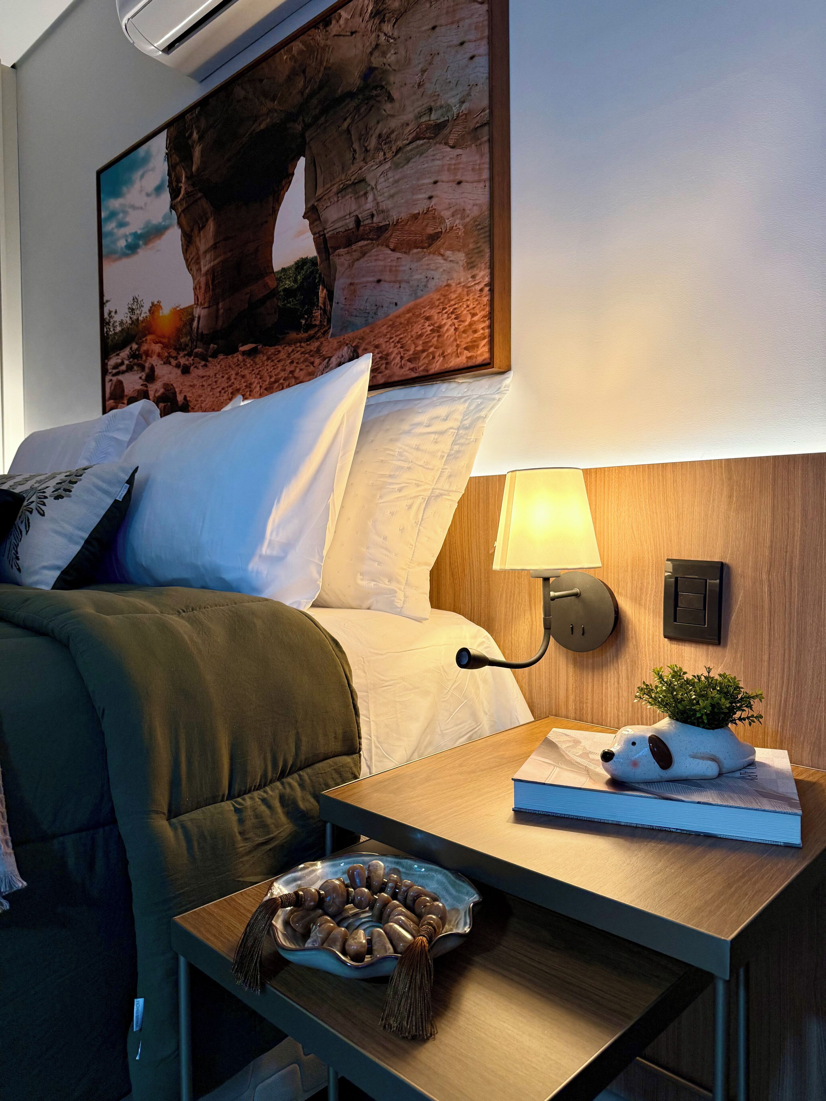 Quarto de hotel bem arrumado, com cama, mesa de cabeceira, abajur e interruptor na parede.