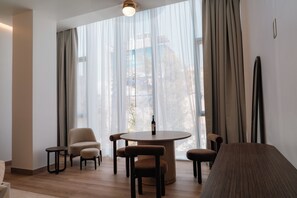 Quarto moderno com mesa redonda, quatro cadeiras, um sofá e uma janela grande com cortinas leves.