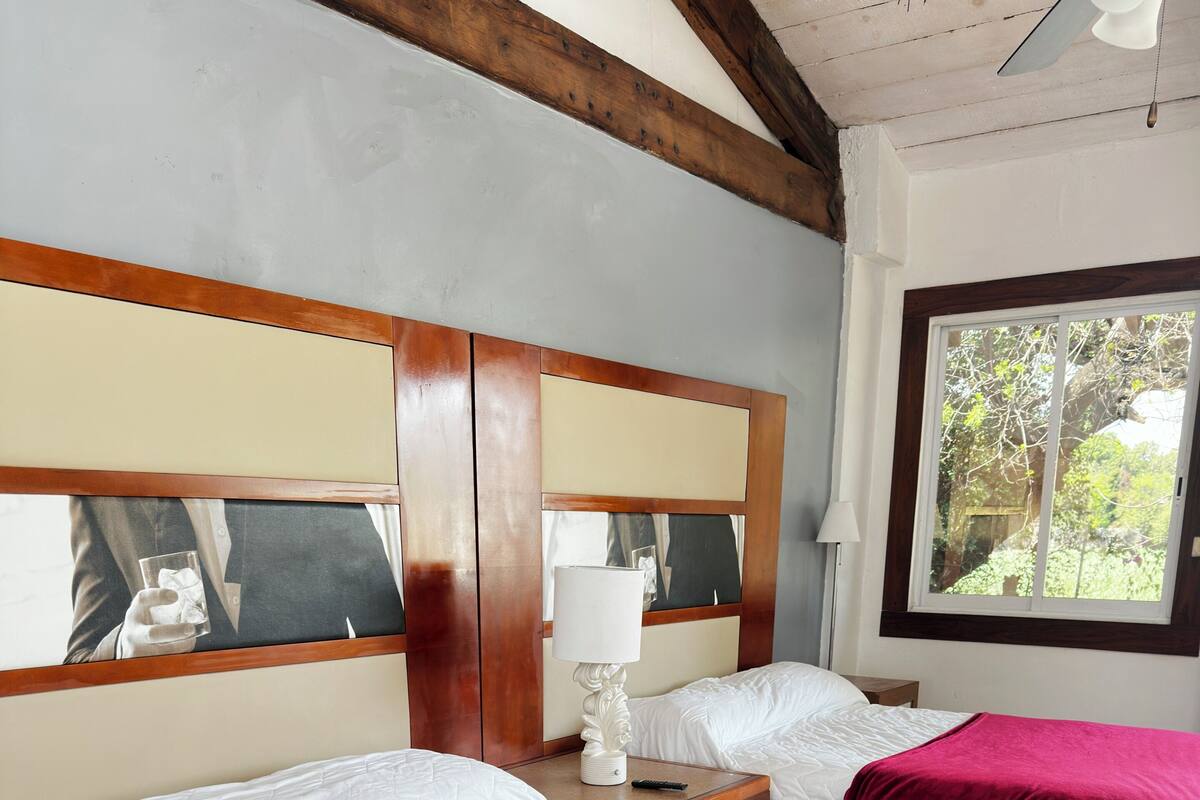 Quarto de hotel com duas camas, uma cabeceira de madeira, uma mesinha de cabeceira com abajur e janela com vista para árvores.