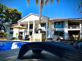 Uma área de piscina com espreguiçadeiras, uma estátua e um edifício com colunas e telhado de telhas vermelhas.