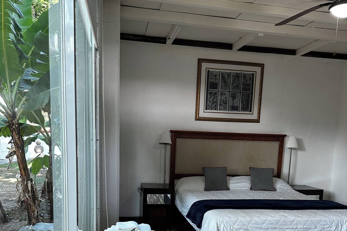 Quarto com cama, mesas de cabeceira, uma pequena mesa com toalhas, ventoinha no teto e um quadro na parede.