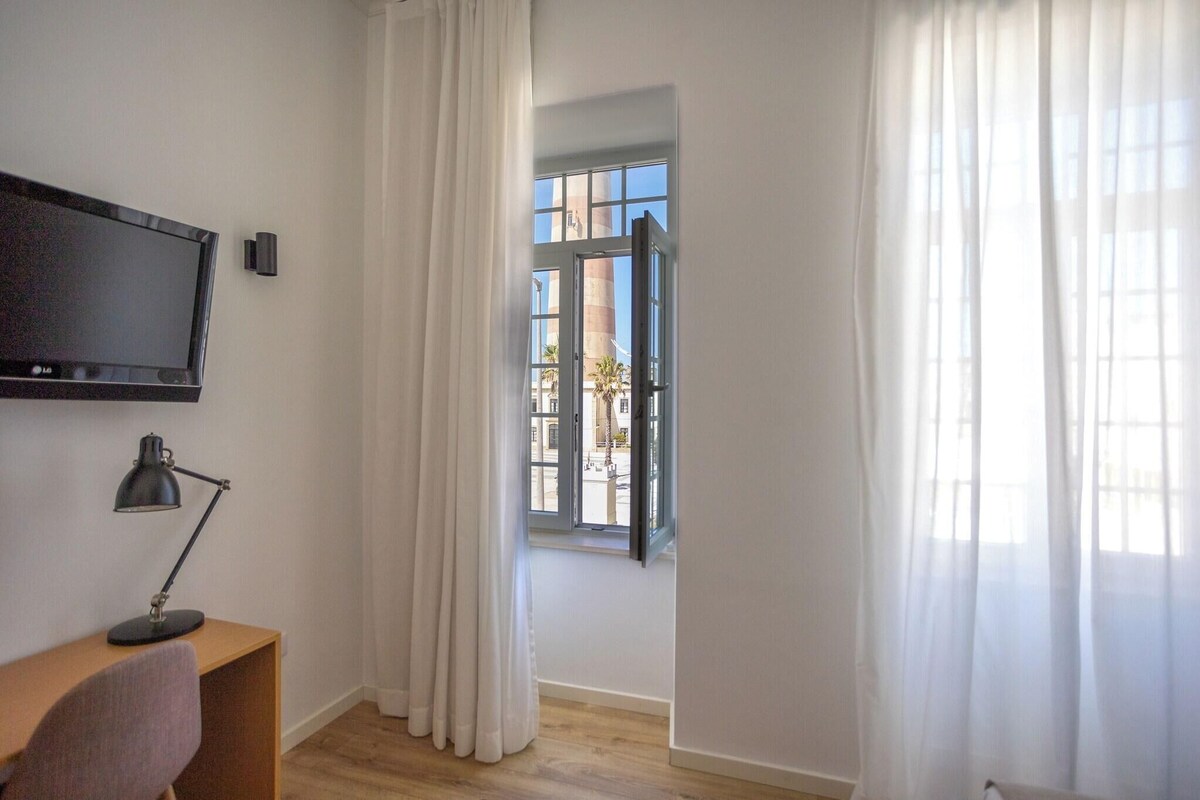 Quarto com televisão na parede, escrivaninha com luminária e vista pela janela com cortinas.