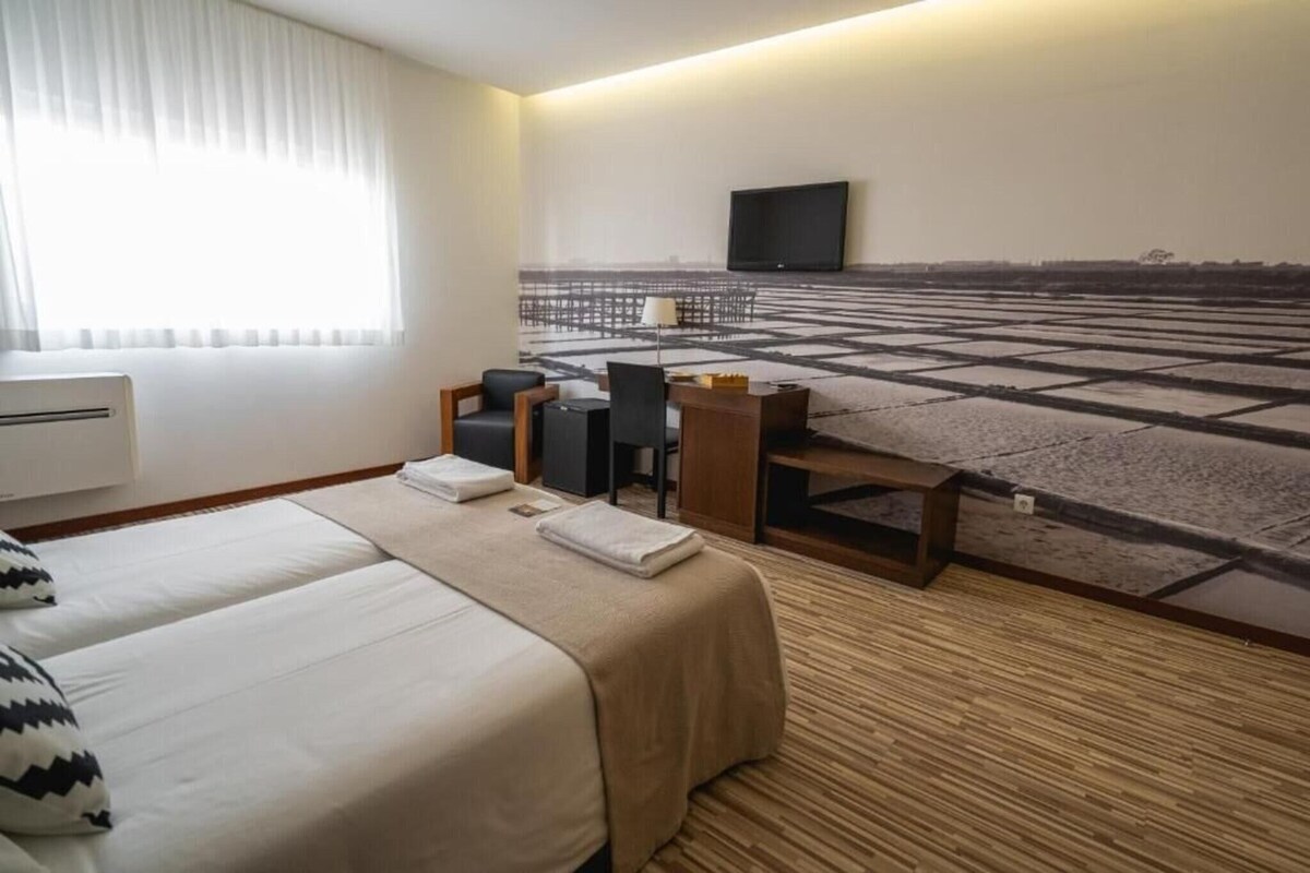 Quarto de hotel com cama, escrivaninha, cadeira, televisão e tapete estampado.