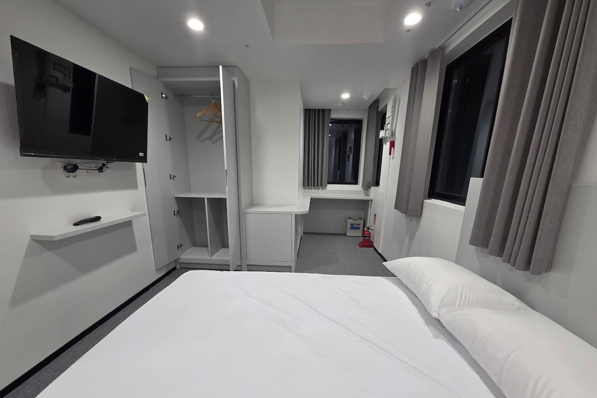 Quarto de hotel com cama, televisão na parede, guarda-roupa e pequena área de trabalho.