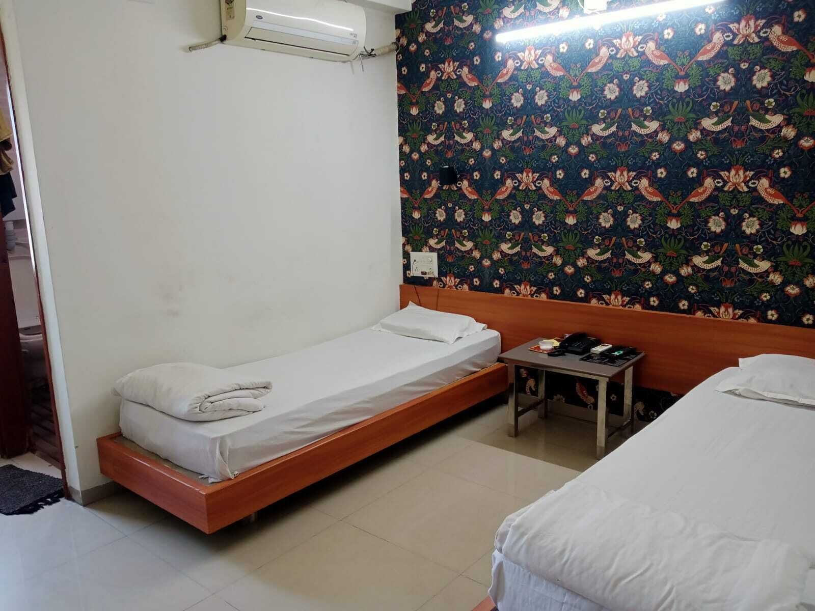 Uma cama de solteiro com estrutura de madeira, uma mesa de cabeceira com telefone e um ar condicionado fixado na parede.