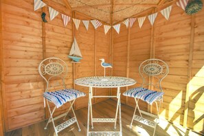 Uma cabana de madeira com uma mesa redonda e duas cadeiras, uma decoração náutica de um veleiro pendurado e uma figura de pássaro.