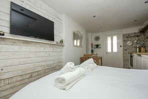 Quarto com televisão fixada em parede de madeira, cama com toalhas dobradas, área de refeições com cadeiras e cozinha com fogão e utensílios.