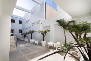 Uma área de pátio moderna ao ar livre, com mobiliário branco, plantas em vasos e uma escadaria que leva a um andar superior.