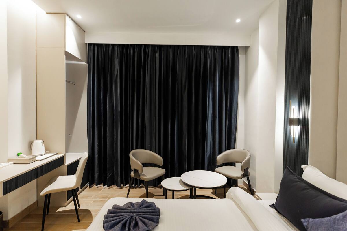 Quarto de hotel moderno com uma cama, duas cadeiras, uma mesinha, uma escrivaninha e uma janela grande com cortinas.