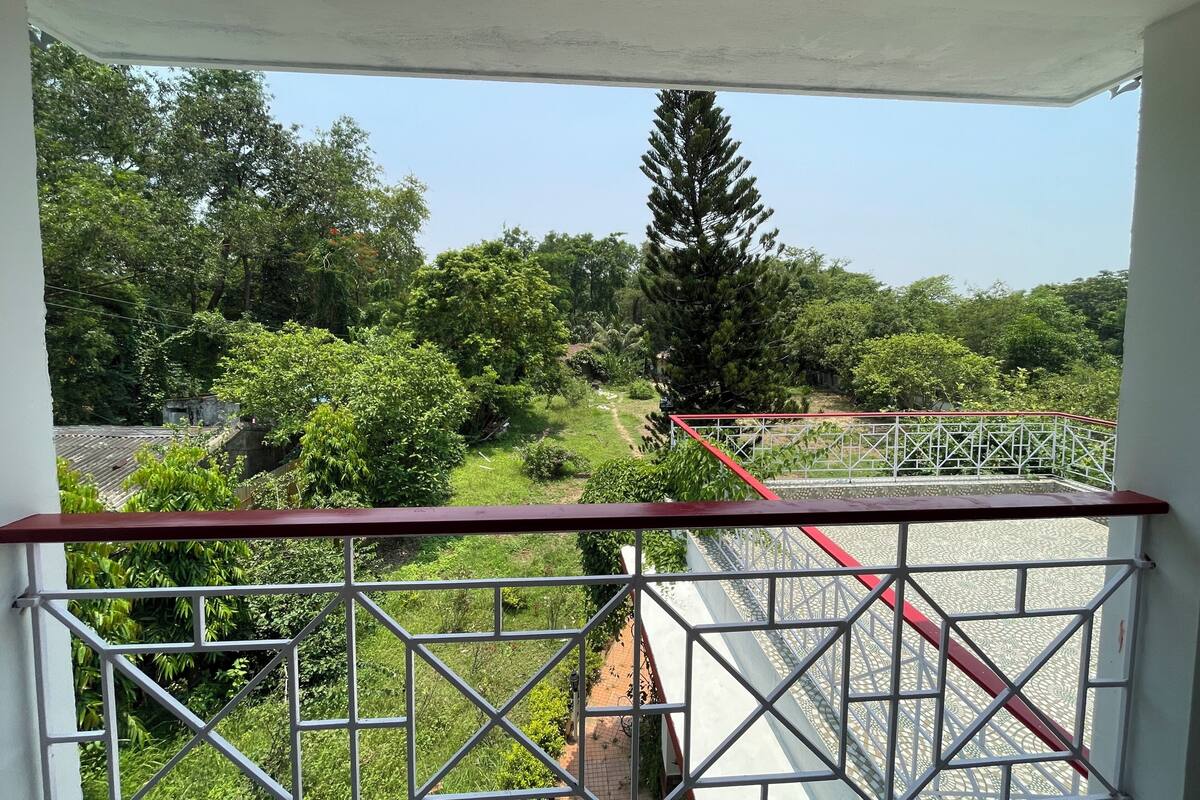 Uma varanda com grade de metal, vista de um jardim com árvores e um caminho, e céu limpo.