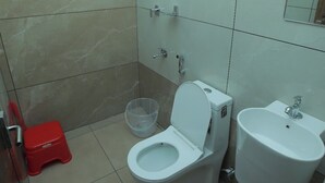 Casa de banho com sanita, lavatório, chuveiro e um banquinho vermelho.