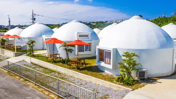 Fileira de edifícios em forma de cúpula, com mesas de piquenique e guarda-sóis vermelhos, cercada por vegetação.